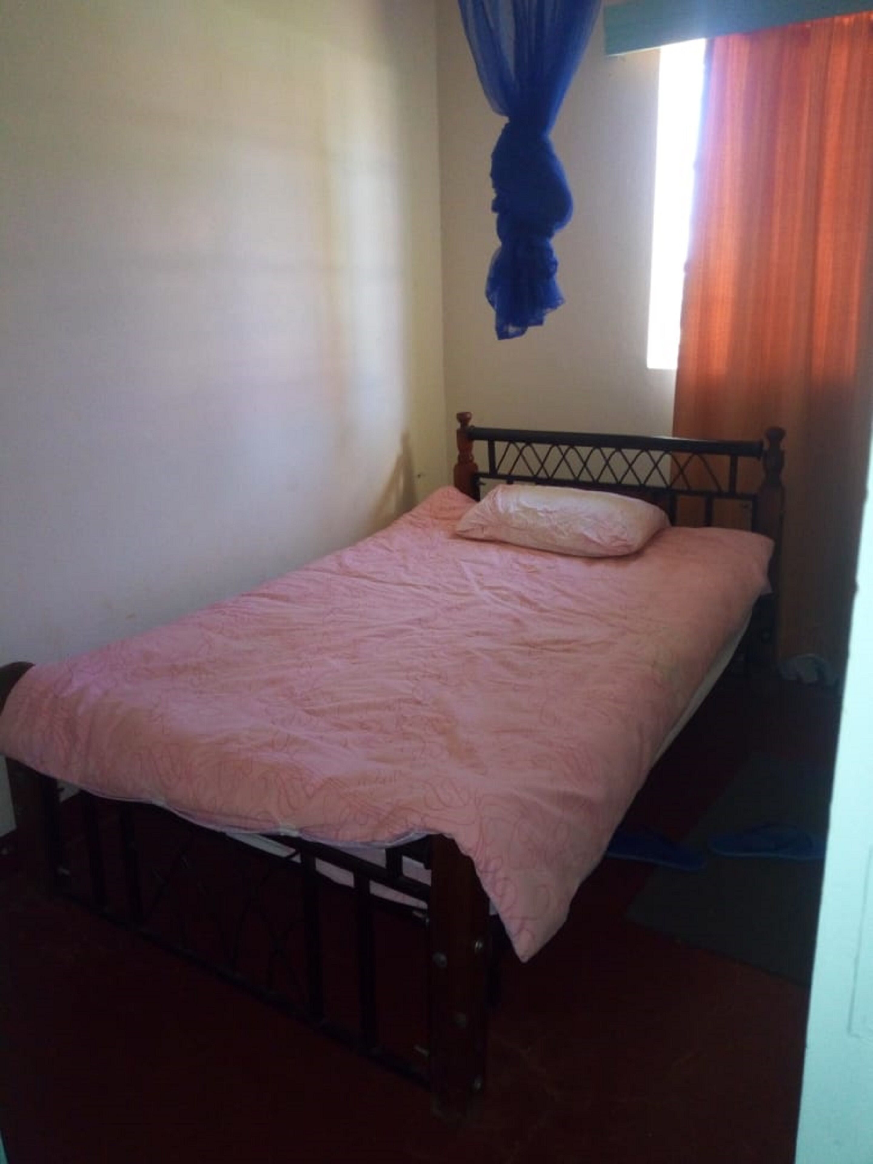 Uma cama de solteiro com estrutura de madeira e colcha rosa, uma janela com cortinas laranja e um mosquiteiro azul.