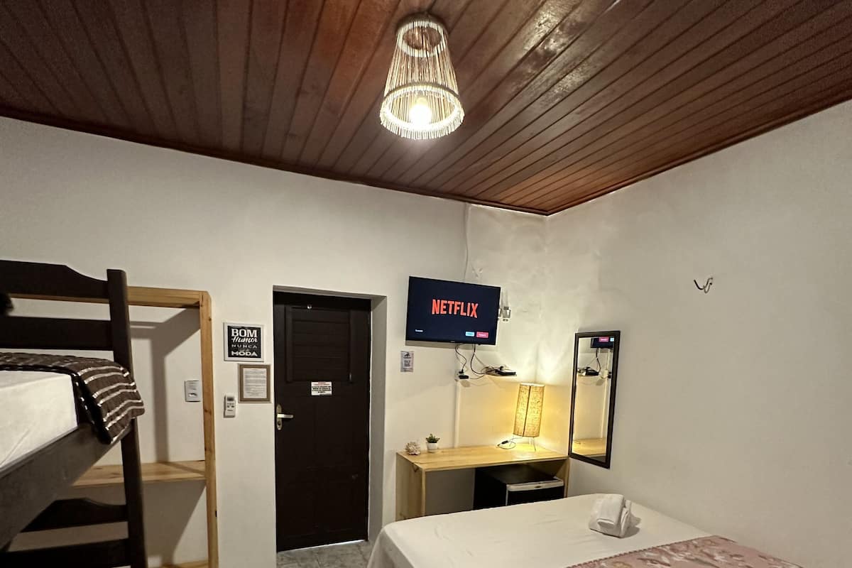 Quarto com beliches, uma escrivaninha com computador, uma televisão exibindo 'NETFLIX' e um espelho na parede.