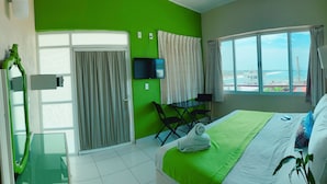 Quarto de hotel com parede verde, cama com colcha verde, televisão na parede, escrivaninha com cadeira e vista da cidade pela janela.