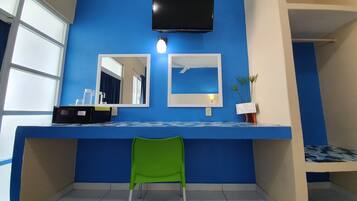 Quarto com parede azul destacada, TV de tela plana acima de um balcão, duas espelhos, uma cadeira verde e um pequeno vaso com planta.