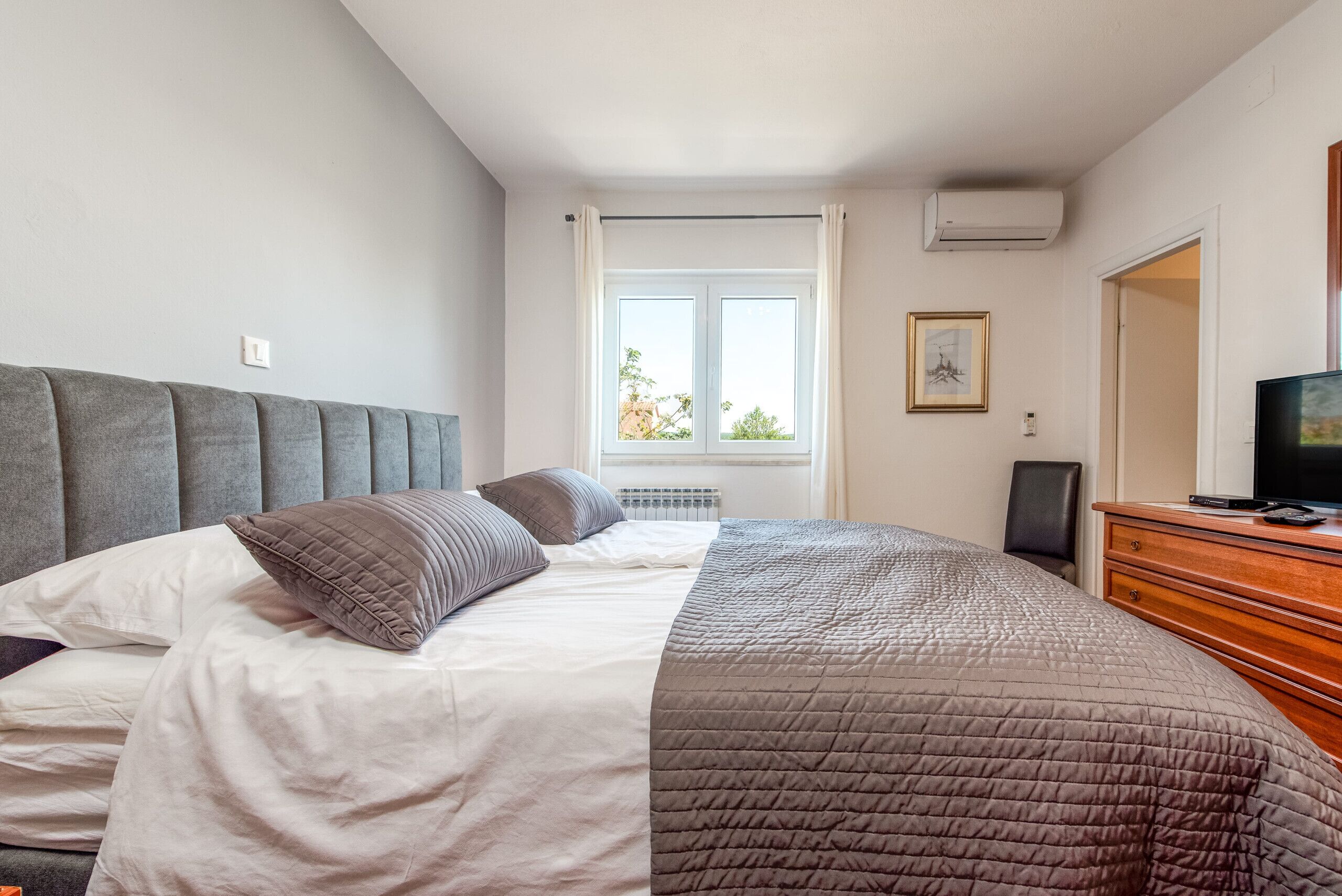 Quarto com cama, penteadeira, televisão, janela com cortinas e aparelho de ar condicionado.