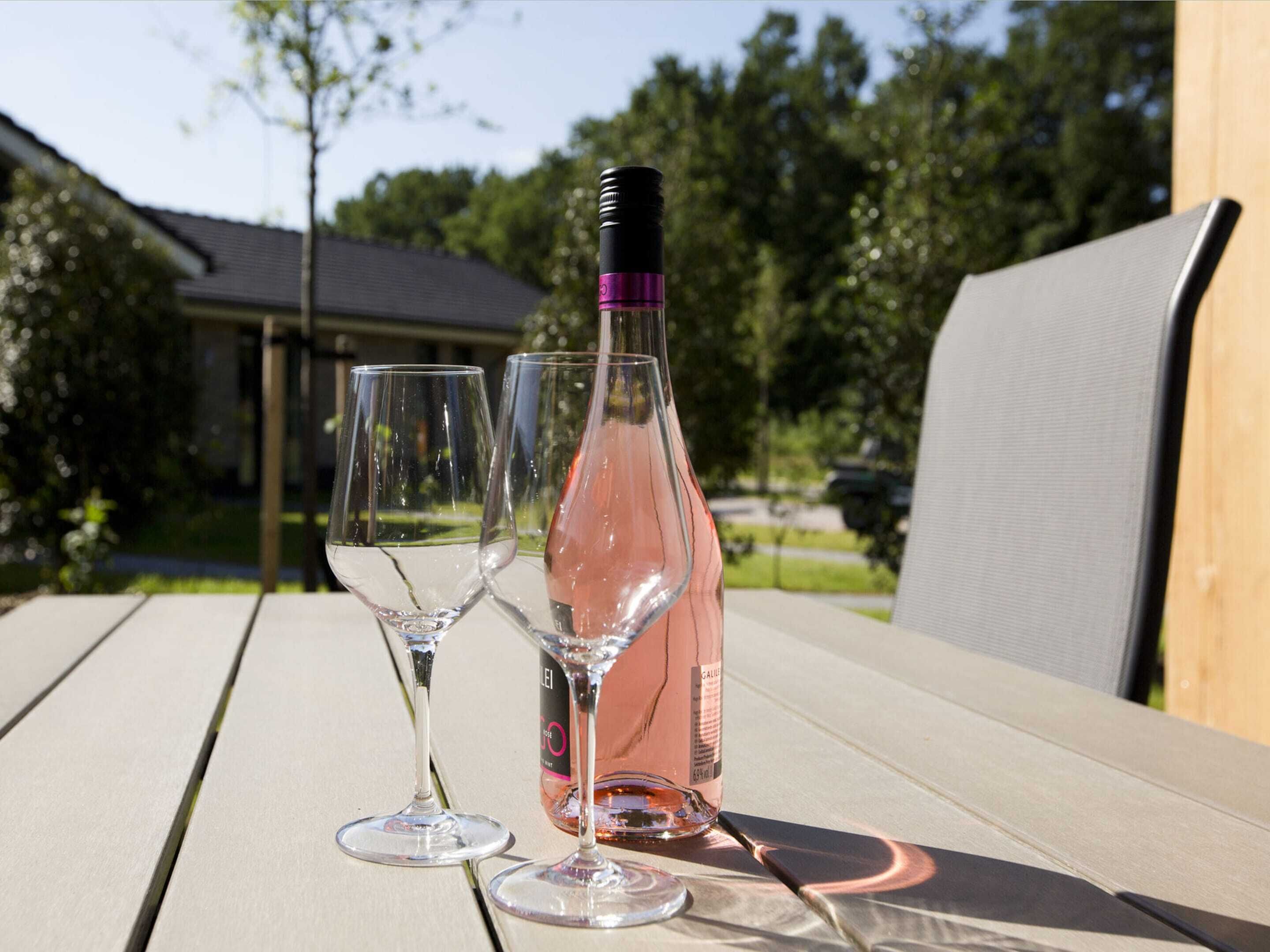 Uma garrafa de vinho rosé e duas taças sobre uma mesa de madeira ao ar livre.