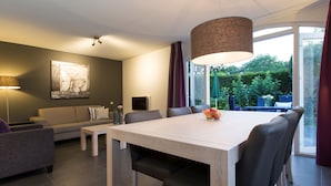 Uma sala de estar moderna com uma mesa de jantar, sofá e cadeiras. Há uma televisão, um quadro e uma janela com vista para o verde.