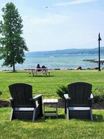 Uma área gramada com duas cadeiras Adirondack, uma mesa de piquenique e vista para um lago com barcos e uma linha de costa distante.
