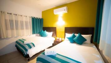 Quarto de hotel com duas camas, uma parede de destaque amarela, um ar condicionado e uma mesa de cabeceira com abajur.