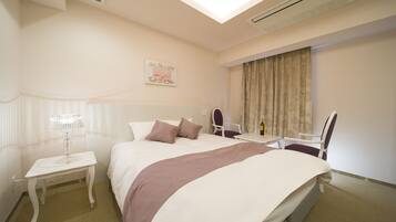 Quarto de hotel com cama, escrivaninha, cadeira, luminária e janela com cortinas.