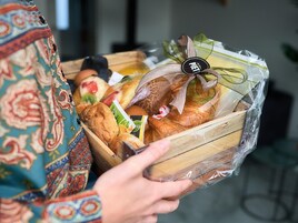 Uma pessoa segurando uma cesta de madeira cheia de pães e doces variados, embalados em plástico transparente com uma fita e uma etiqueta.