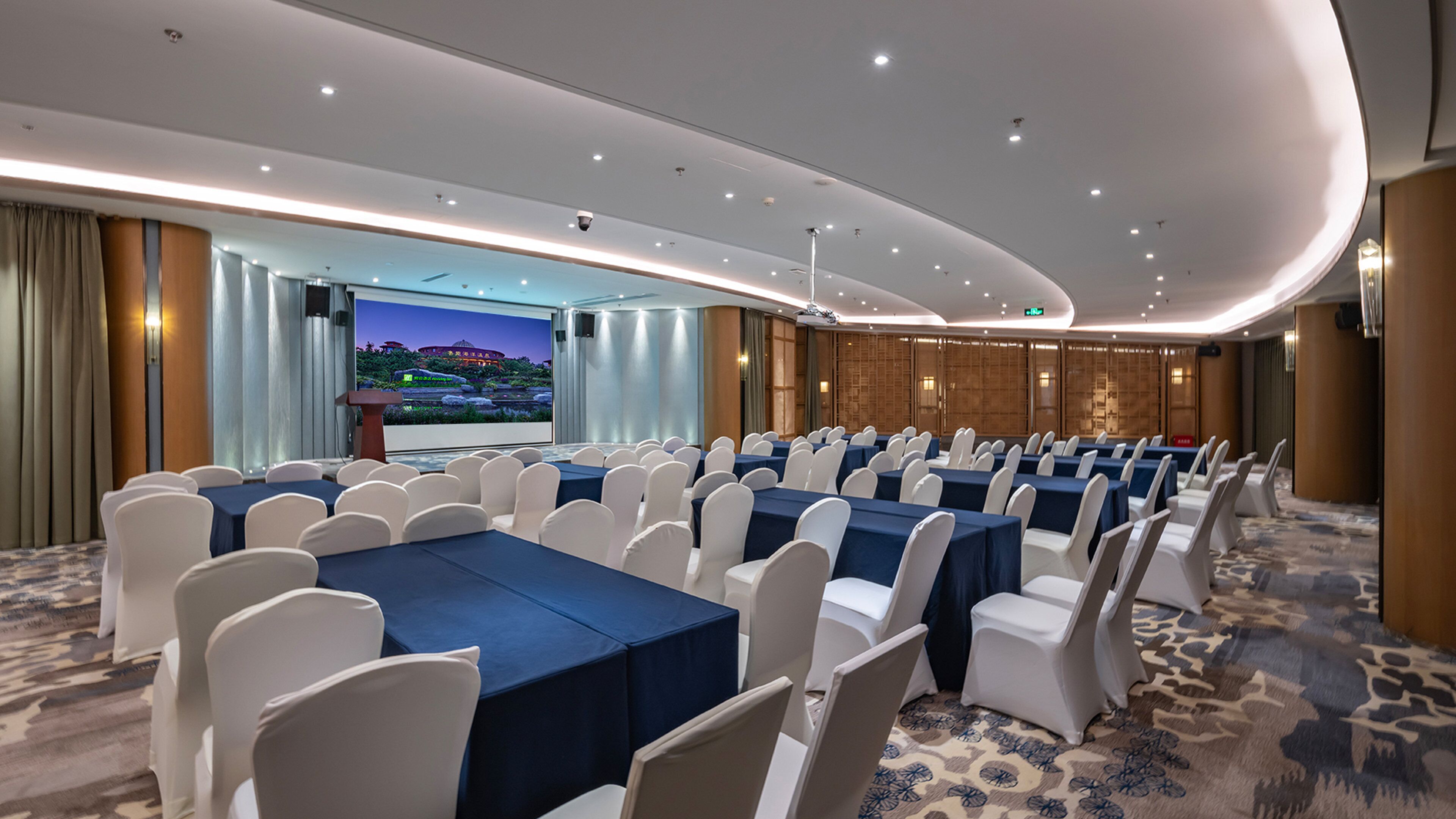 Sala de conferência com mesas e cadeiras dispostas em fileiras, um púlpito e uma tela grande exibindo uma vista panorâmica.
