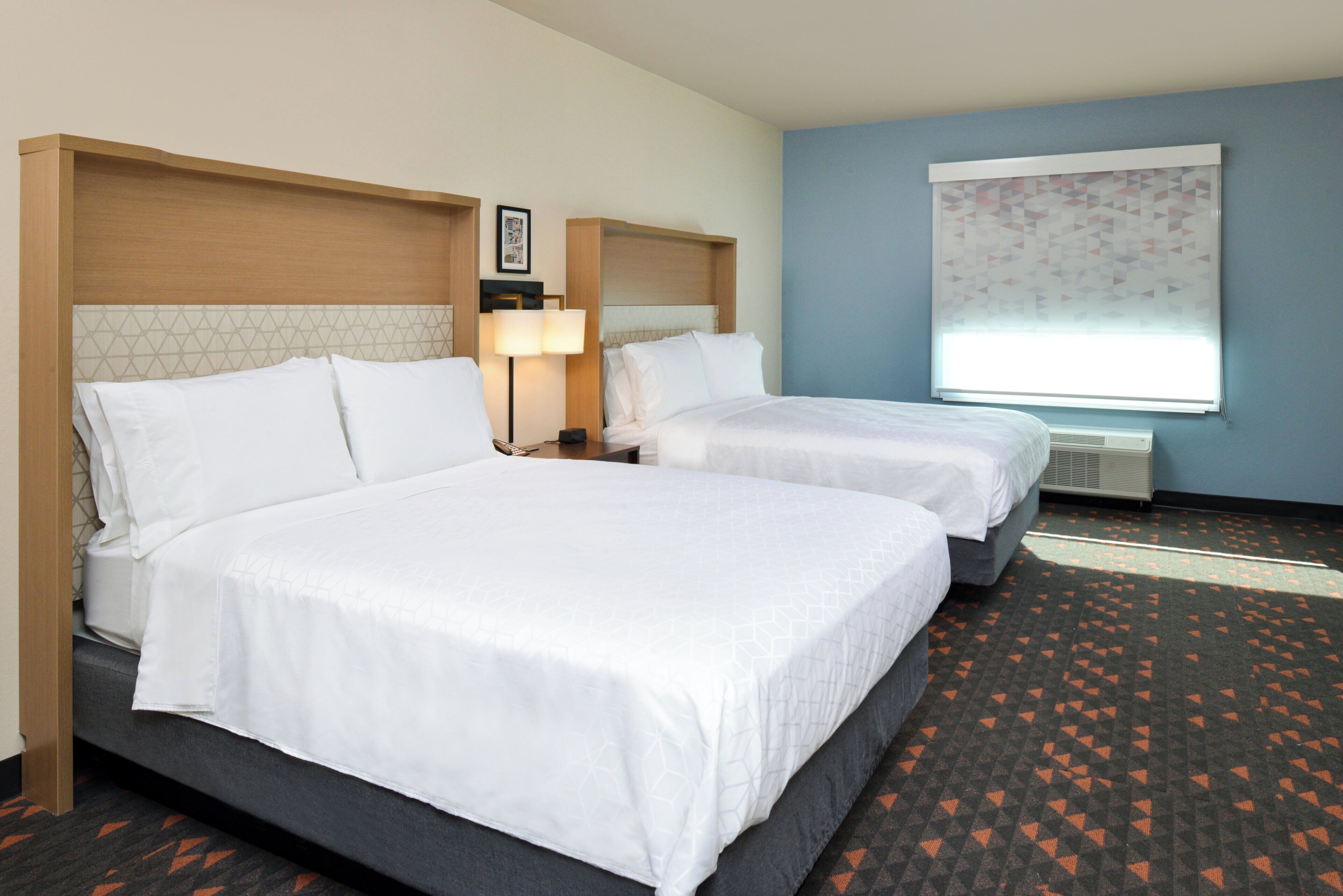 Quarto de hotel com duas camas, uma mesa de cabeceira com abajur, uma janela com persianas e um tapete estampado.
