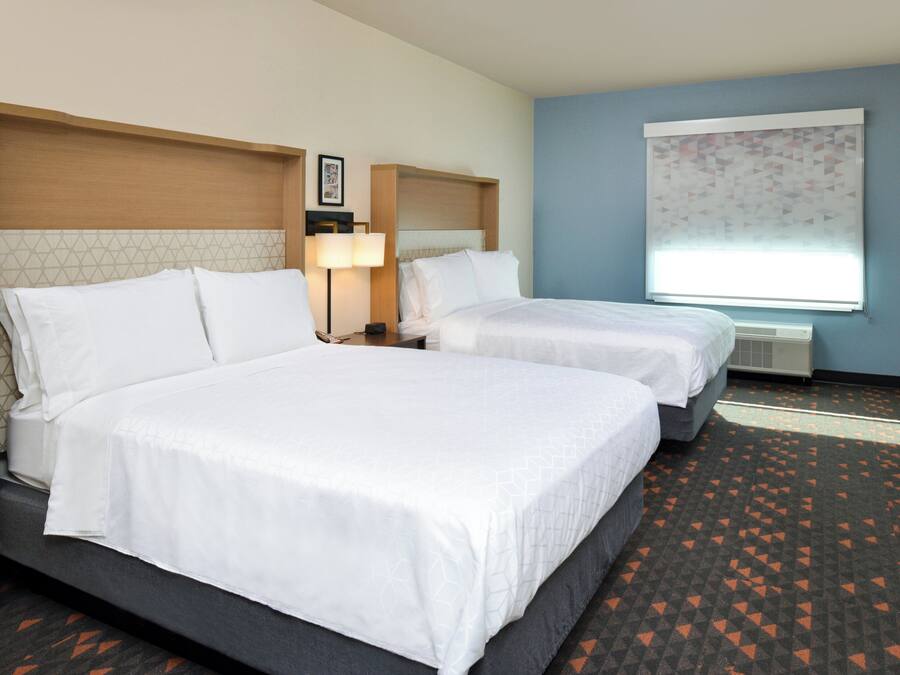 Quarto de hotel com duas camas, uma mesa de cabeceira com abajur, uma janela com persianas e um tapete estampado.