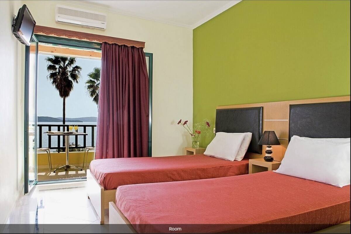 Quarto de hotel com duas camas, uma varanda com vista para palmeiras e uma pequena mesa com flores.