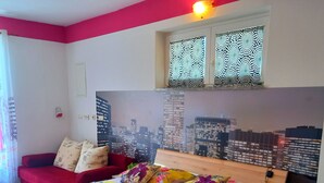Quarto com um mural da paisagem urbana na parede, uma cama com roupa de cama floral, um sofá vermelho e uma mesa de cabeceira com uma planta.