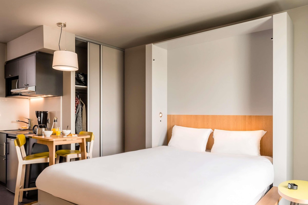 Quarto de hotel com cama, escrivaninha, cadeira, mesa pequena e luminária pendente.