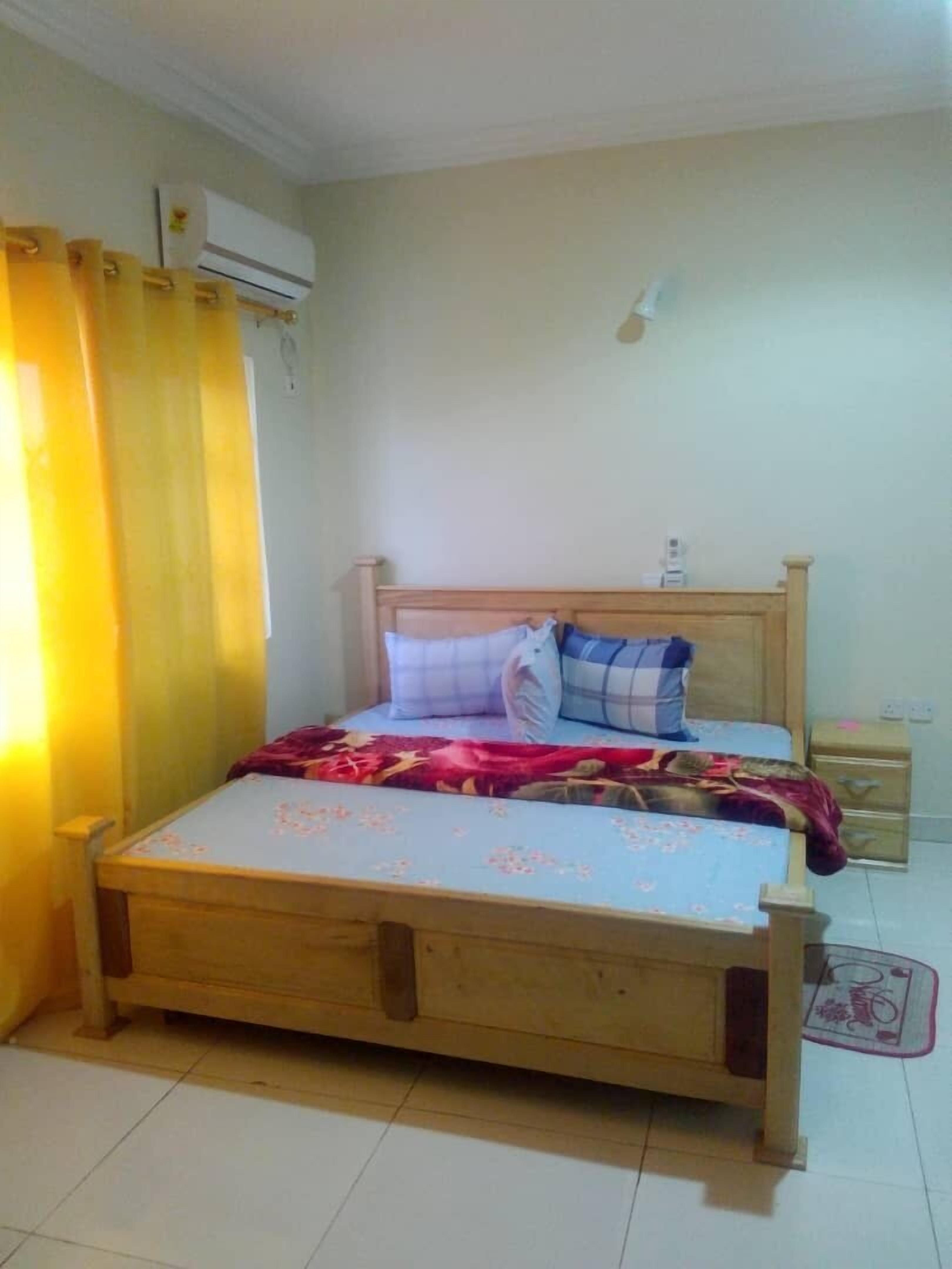 Quarto individual com uma cama de madeira, colcha floral, mesa de cabeceira com luminária e unidade de ar condicionado na parede.