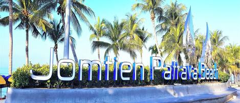 Um letreiro com a inscrição 'Jomien Pattaya Beach', rodeado por palmeiras e um céu claro.