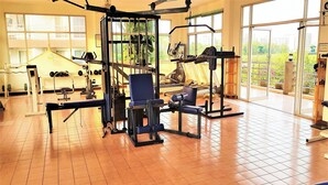 Academia com diversos equipamentos de exercício, amplas janelas e piso em azulejo.