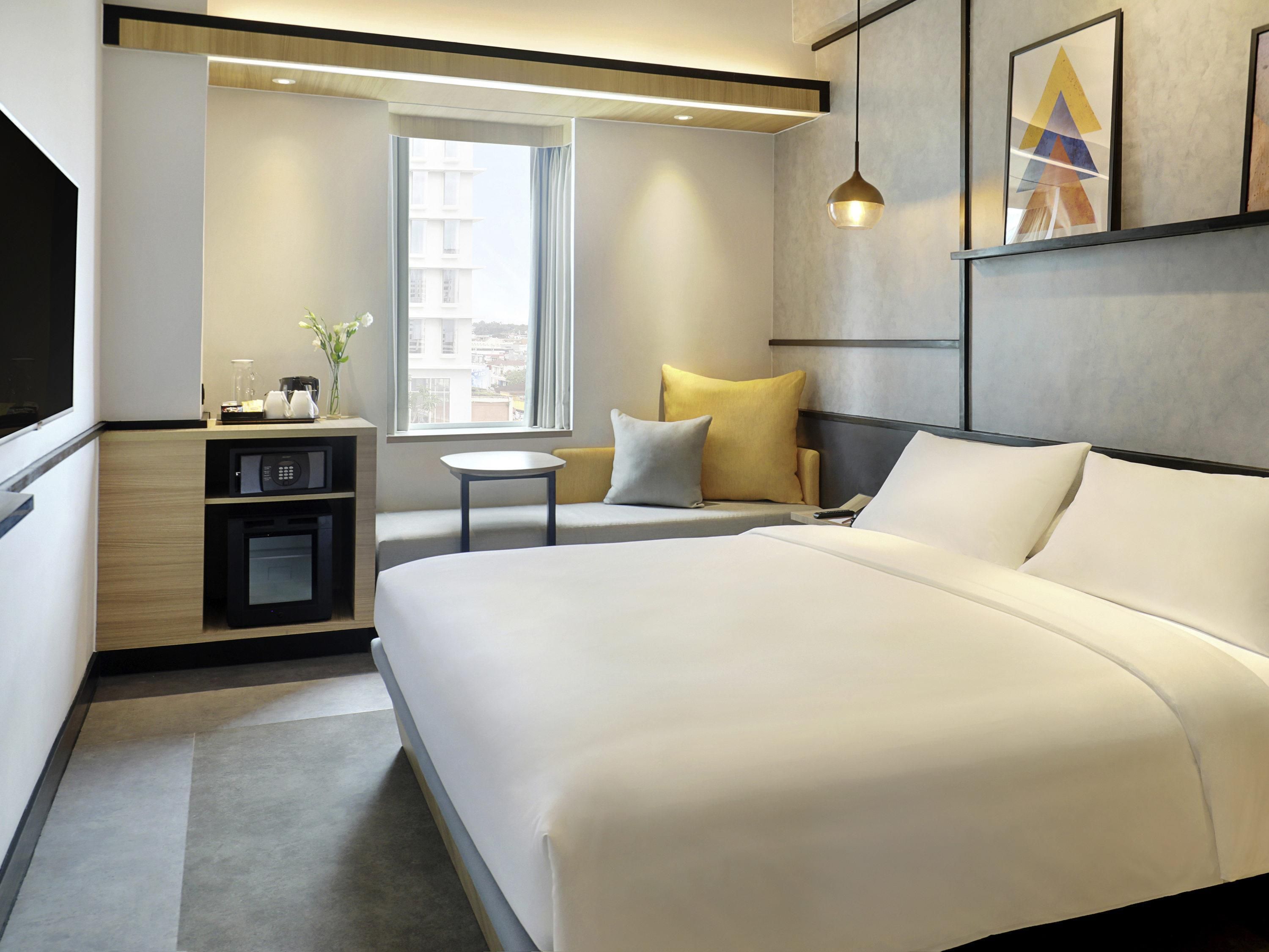 Quarto de hotel moderno com uma cama grande, uma mesinha, micro-ondas e janela com vista para a cidade.