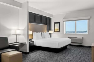 Quarto de hotel com uma cama grande, uma escrivaninha com luminária, uma cadeira, uma janela com vista para a cidade e um aparelho de ar condicionado.