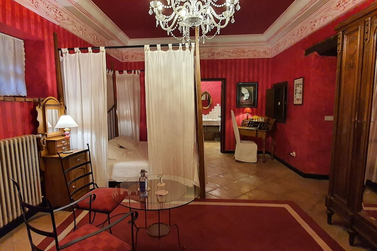 Quarto com parede listrada em vermelho e branco, um lustre, uma cama com cortinas brancas, cômoda de madeira e um radiador.
