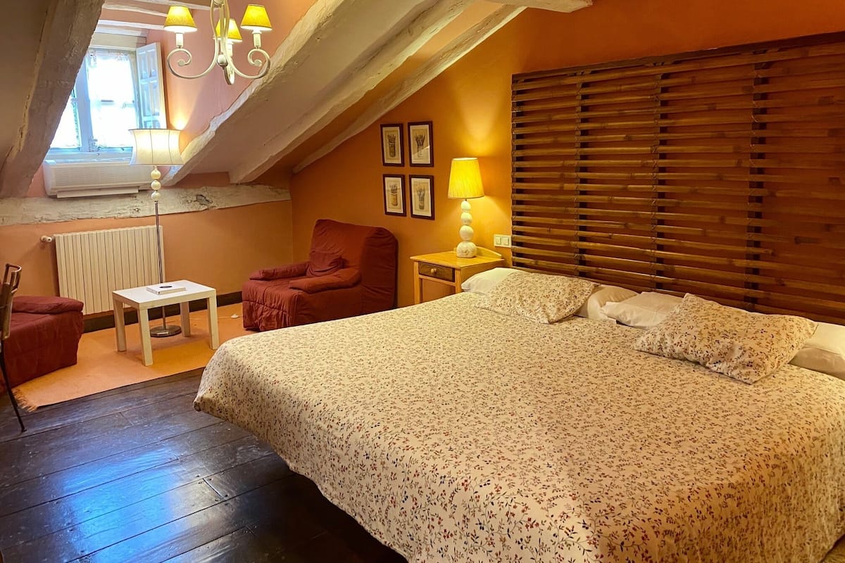 Quarto com cama grande, uma cadeira, mesa pequena, abajur e janela com persianas.