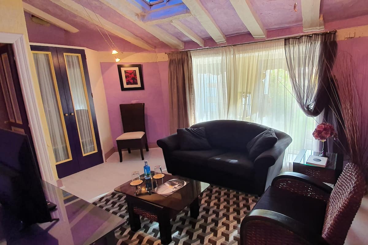 Quarto com sofá de couro preto, uma mesa pequena com um copo, uma garrafa e um quadro emoldurado. Há uma cadeira, um radiador e uma janela com cortinas.