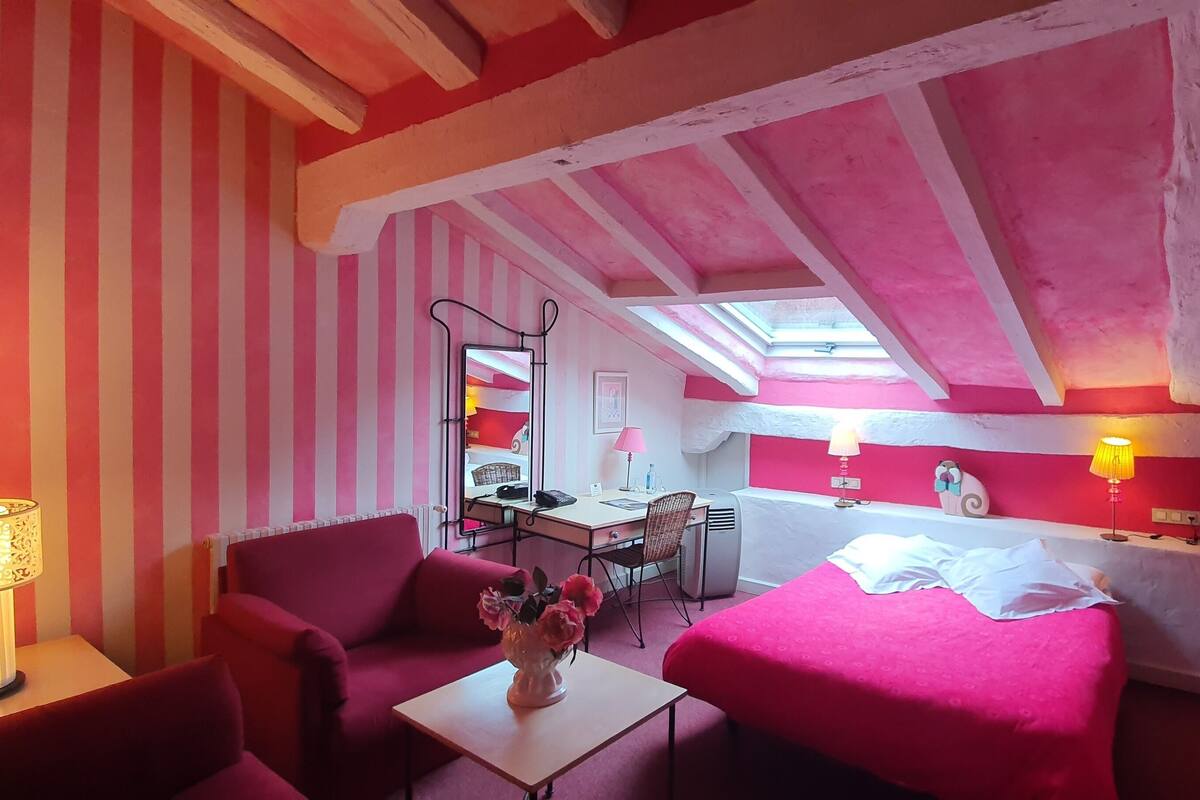 Um quarto com uma parede listrada em vermelho e branco, um sofá vermelho, uma pequena mesa com um abajur, uma cama com colcha rosa e um claraboia sobre uma escrivaninha com cadeira e abajur.