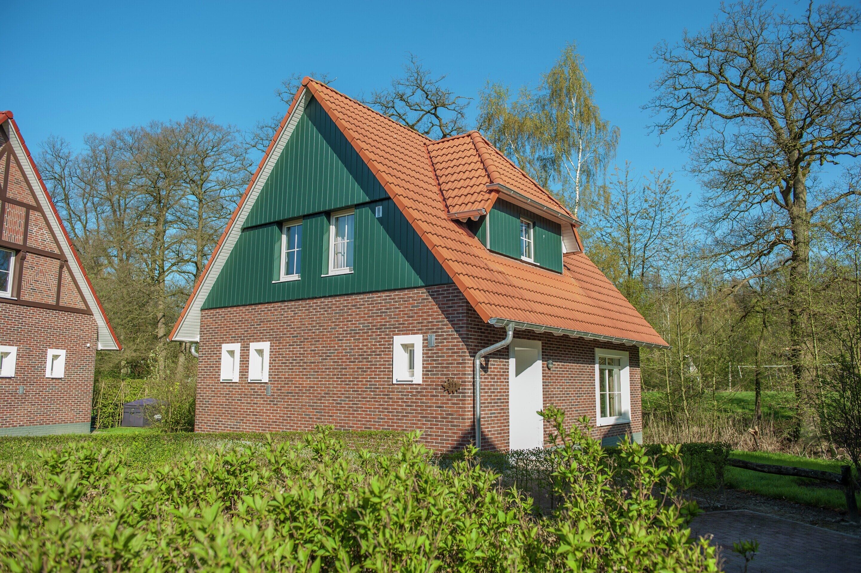 Uma casa de dois andares com fachada verde e telhado de telhas vermelhas, rodeada por vegetação e árvores.