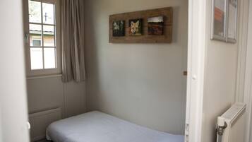 Quarto pequeno com uma cama de solteiro, janela, radiador e três quadros na parede.