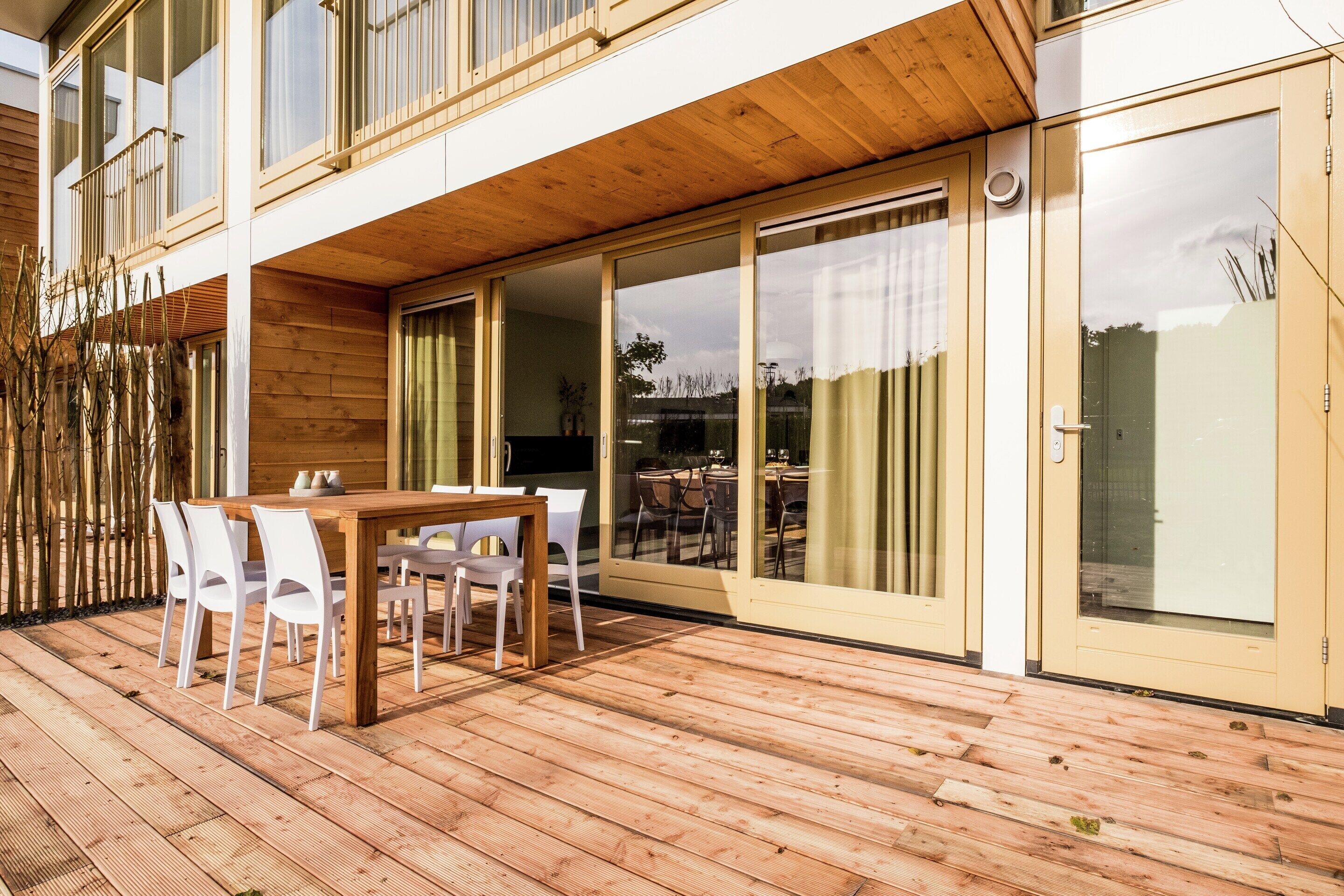 Um deck de madeira com mesa e cadeiras, uma porta de correr de vidro que dá para uma área de refeições ao ar livre e um corrimão de varanda em madeira.