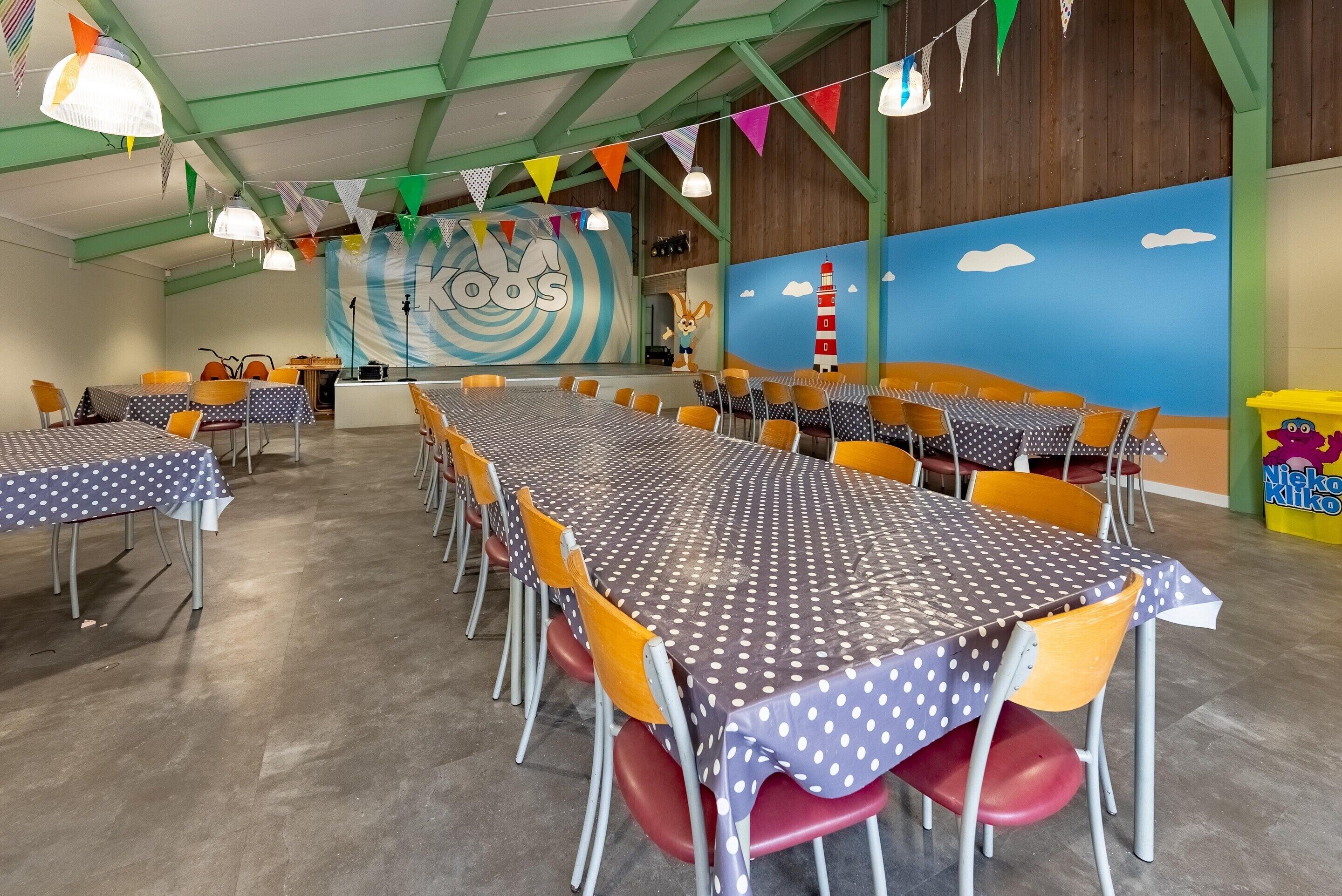 Um espaço amplo e coberto, com várias mesas e cadeiras, palco com painel de fundo e decorações coloridas.