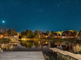 Cena noturna tranquila à beira de um lago, com um píer de madeira que se estende sobre águas calmas, cercado por casas com janelas visíveis e um céu noturno límpido.
