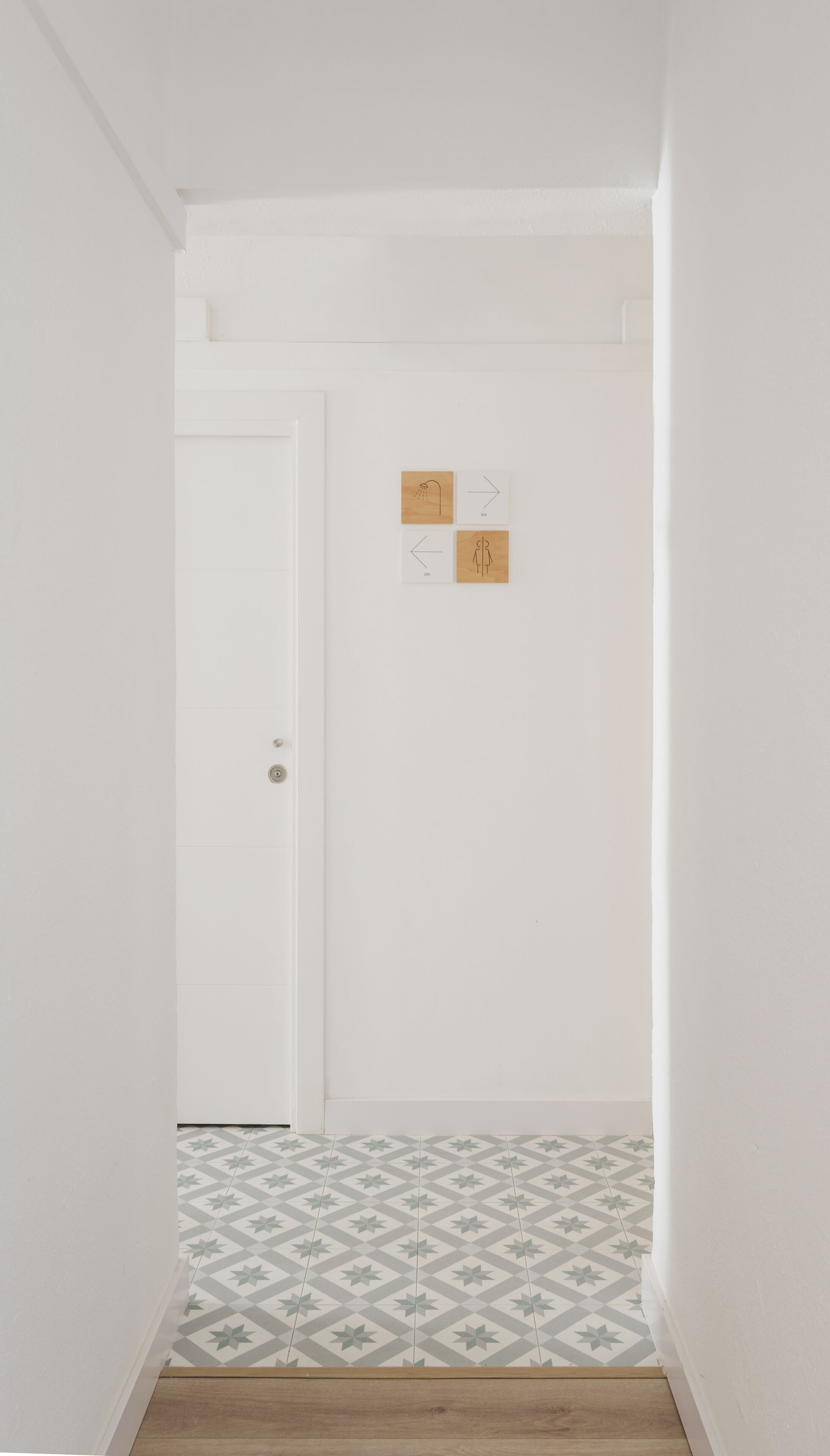 Um corredor com uma porta branca, piso estampado e duas placas fixadas na parede indicando direções e um banheiro.