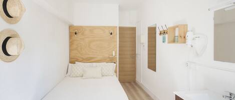Quarto com cabeceira de madeira, cama branca, piso de madeira, banheiro branco com espelho e dois chapéus na parede.