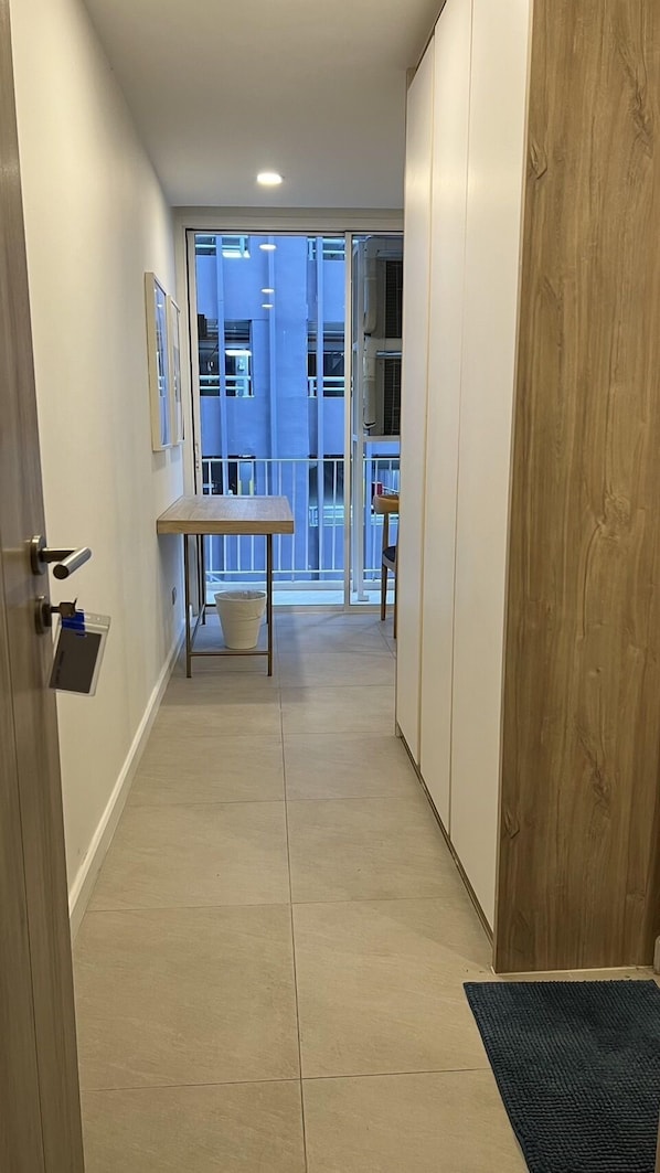 Um corredor com uma porta à esquerda, uma mesa e uma cadeira no centro, e vista de um edifício com varandas através de uma porta de vidro.