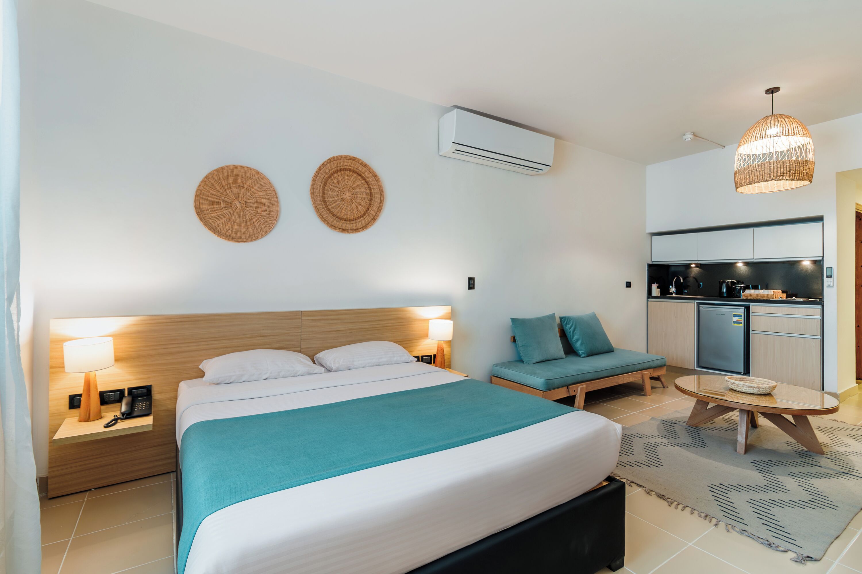 Quarto de hotel moderno com cama grande, mesas de cabeceira, um pequeno sofá, área de cozinha e mesa de jantar.