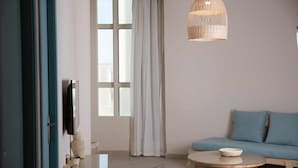 Um quarto com sofá, uma mesa redonda, luminária pendente, janela com cortinas e televisão na parede.