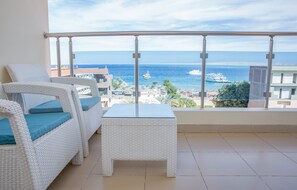 Varanda com mobiliário de vime branco e mesa de vidro, com vista para a área costeira com barcos e edifícios.