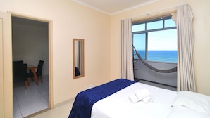 Quarto de hotel com uma cama, vista para o mar através de uma janela, uma rede e um espaço de refeições visível por uma porta aberta.