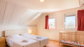 Quarto com duas camas, um berço, piso de madeira, janela com cortinas e radiador.