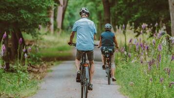 Dois ciclistas em um caminho estreito, cercado por vegetação exuberante e flores roxas.