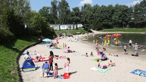 Uma praia com pessoas tomando sol, nadando e brincando. Há barracas, guarda-sóis e um tobogã d'água.