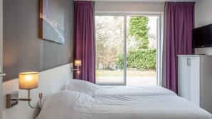 Quarto de hotel com cama, televisão, luminária fixada na parede e janela com cortinas roxas.