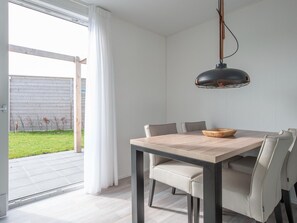 Uma área de refeições com uma mesa de madeira e quatro cadeiras, uma luminária pendente e vista para um jardim através de uma porta deslizante.
