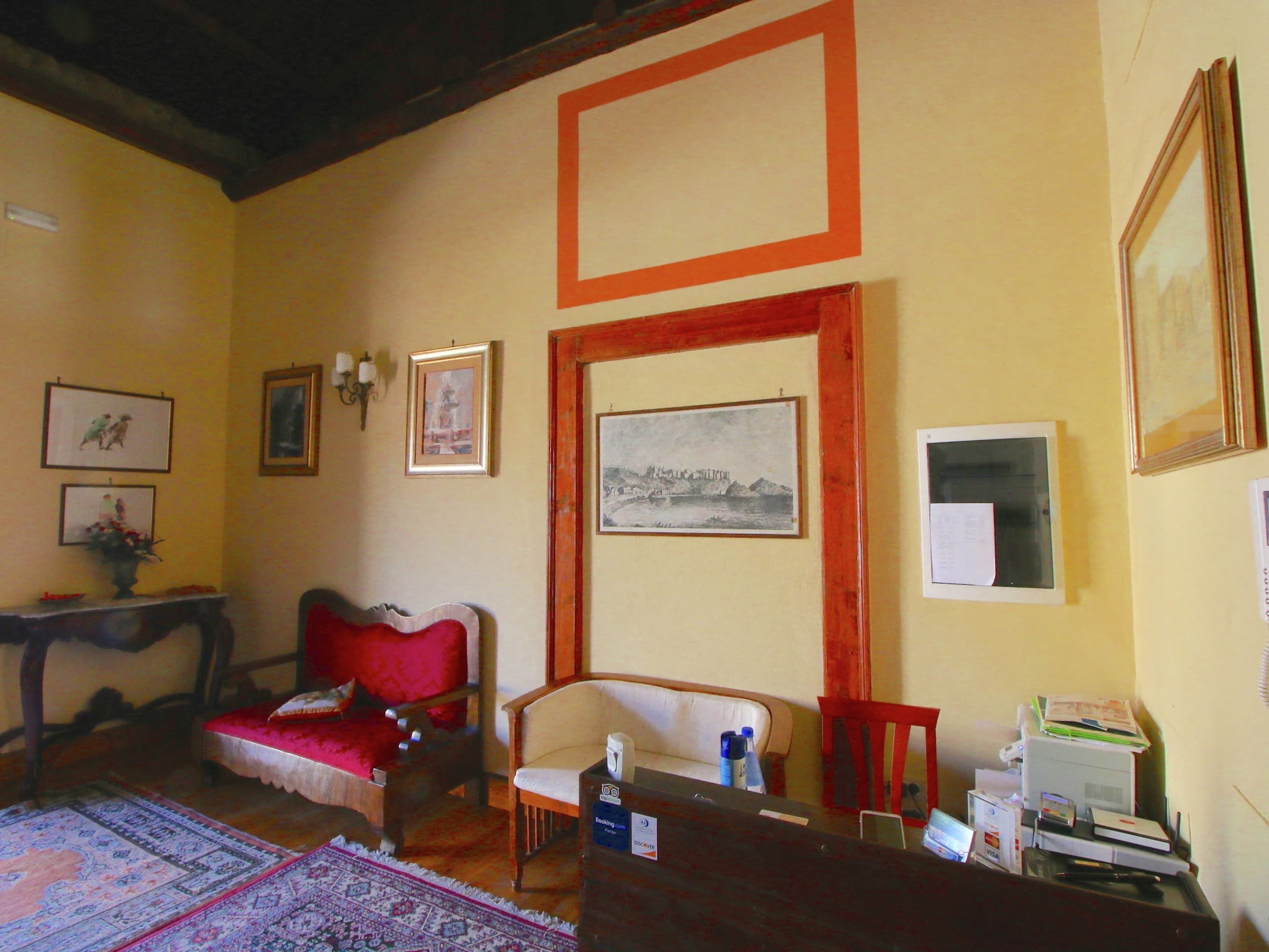 Um quarto acolhedor com um sofá vermelho, uma poltrona branca e uma escrivaninha de madeira com um computador. Há quadros nas paredes e um tapete no chão.