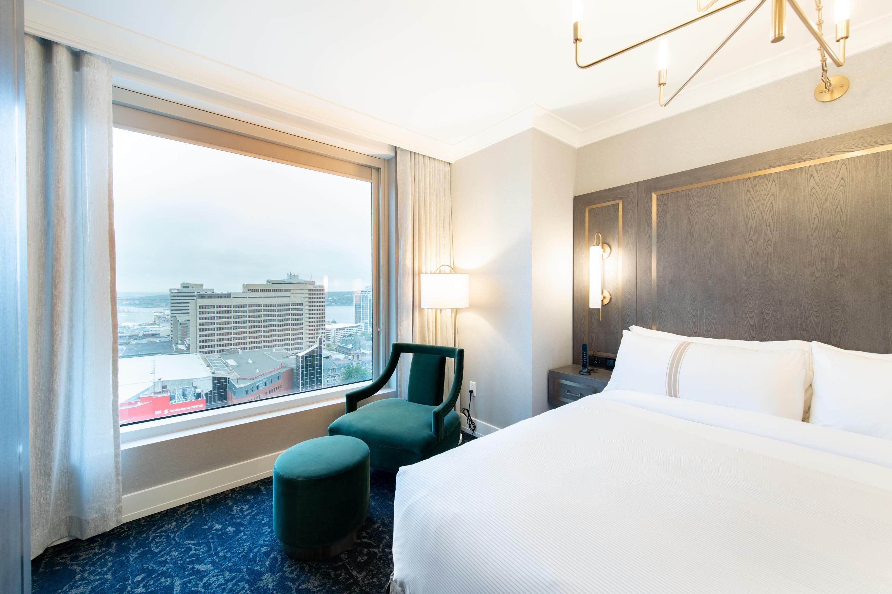 Quarto de hotel com uma janela grande, uma cama, uma cadeira verde e uma mesa de cabeceira com abajur.