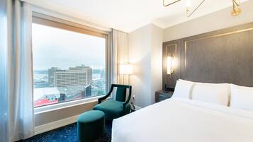 Quarto de hotel com uma janela grande, uma cama, uma cadeira verde e uma mesa de cabeceira com abajur.