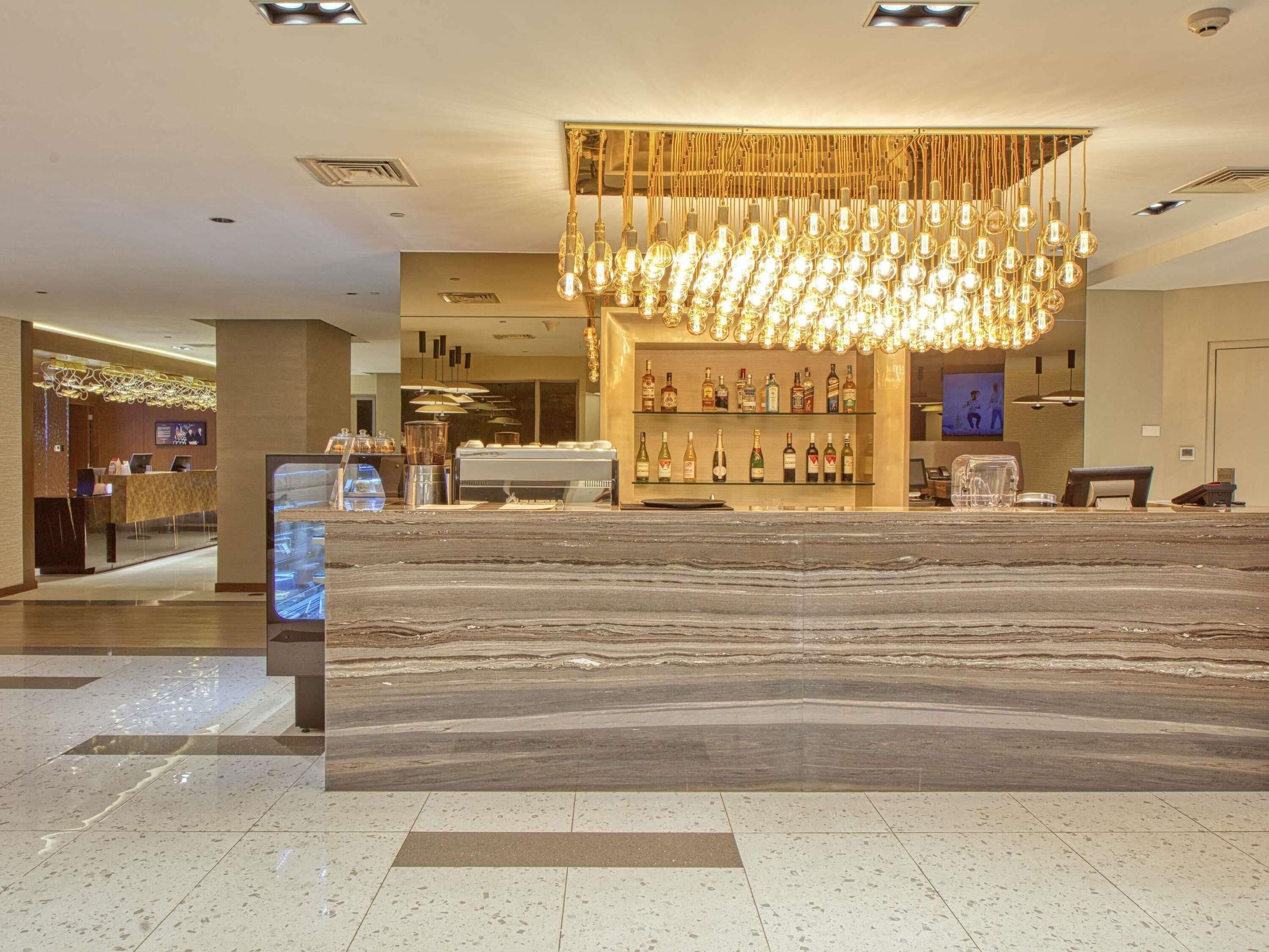 Um saguão de hotel com uma recepção de mármore, área de bar com garrafas de bebidas e um lustre moderno.
