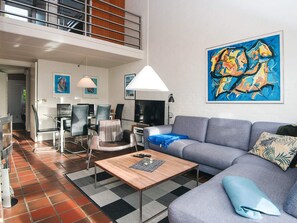 Uma sala de estar moderna com um sofá cinza, uma mesa de centro de madeira e uma televisão. Há um quadro na parede e uma escada que leva a um andar superior.