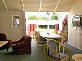 Uma área de refeições com mesa e cadeiras, uma televisão presa na parede e uma janela com cortinas.