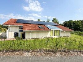 Uma casa térrea com telhado de telhas vermelhas, painéis solares e uma pequena garagem anexa.